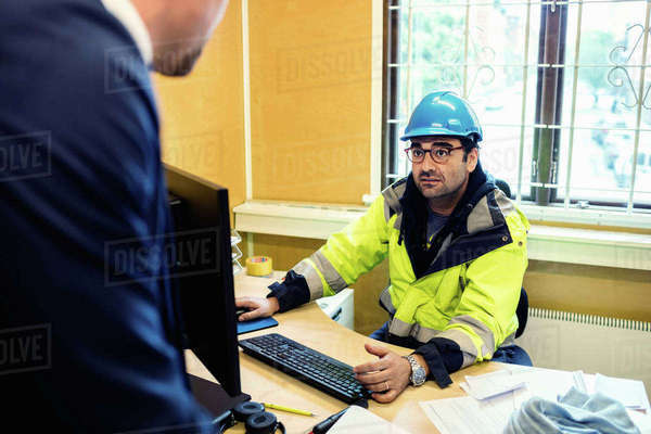 Serious construction worker in protective wear talking to man at desk ...