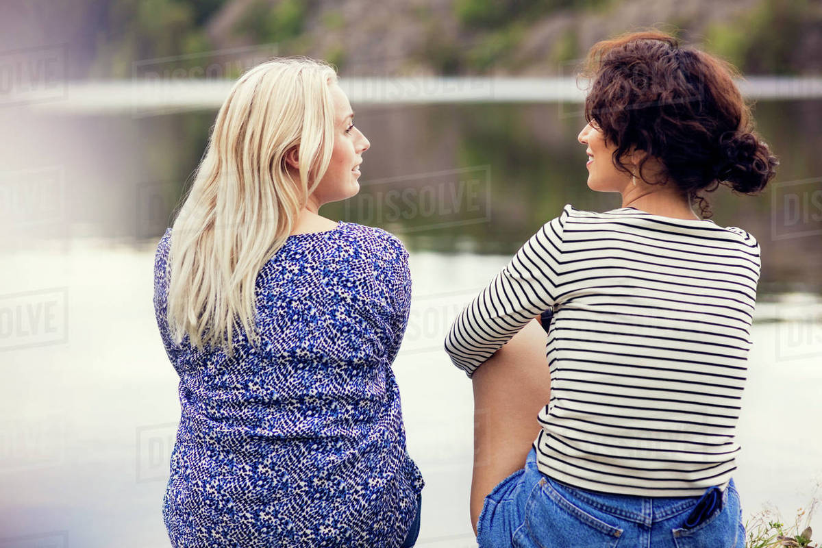 Rear view of friends talking while sitting at lakeshore - Royalty-free ...