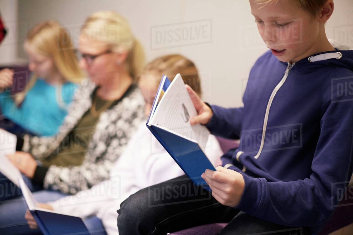 Boy reading book with friends and teacher in background at classroom ...