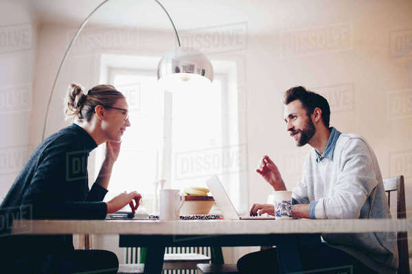 Side view of couple talking while sitting at dining table at home ...
