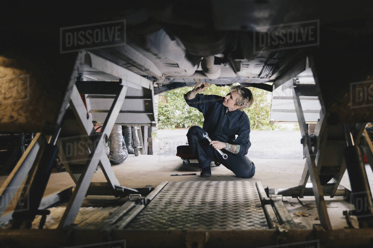 Female mechanic kneeling while examining under car at auto repair shop ...