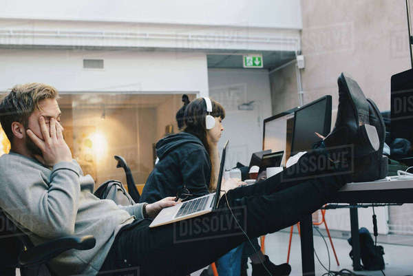 Tired computer programmer sitting with feet up on desk while colleagues ...