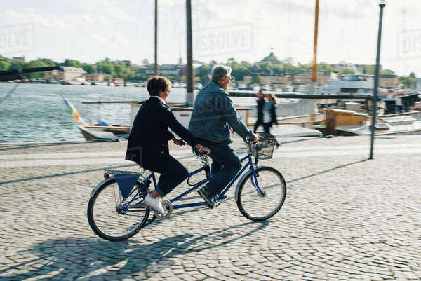 Full length of senior couple riding tandem bike on road in city during ...