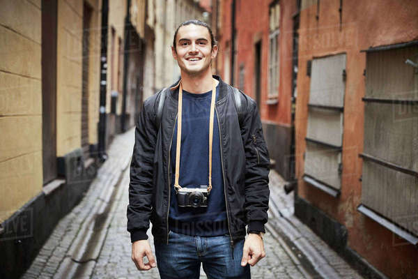 Portrait of smiling man wearing camera standing on cobbled street in ...