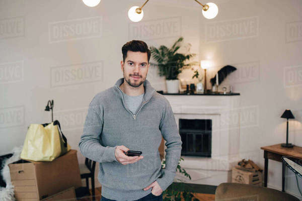 Portrait of smiling man holding mobile phone while standing with hand ...