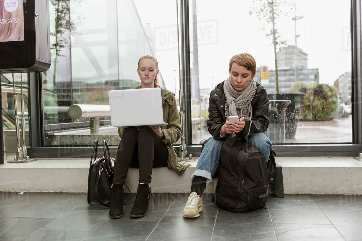 Full length of friends using technologies while waiting at station ...