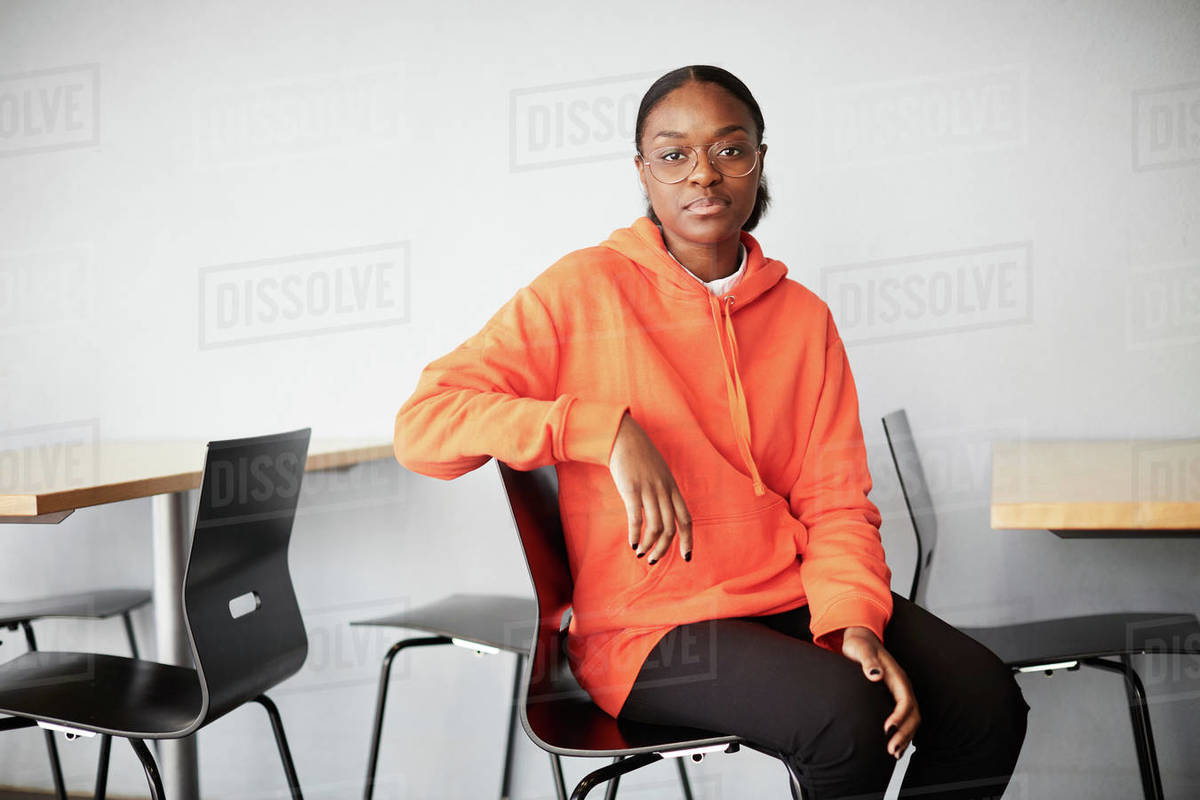 Portrait of young student sitting on chair against wall at university ...