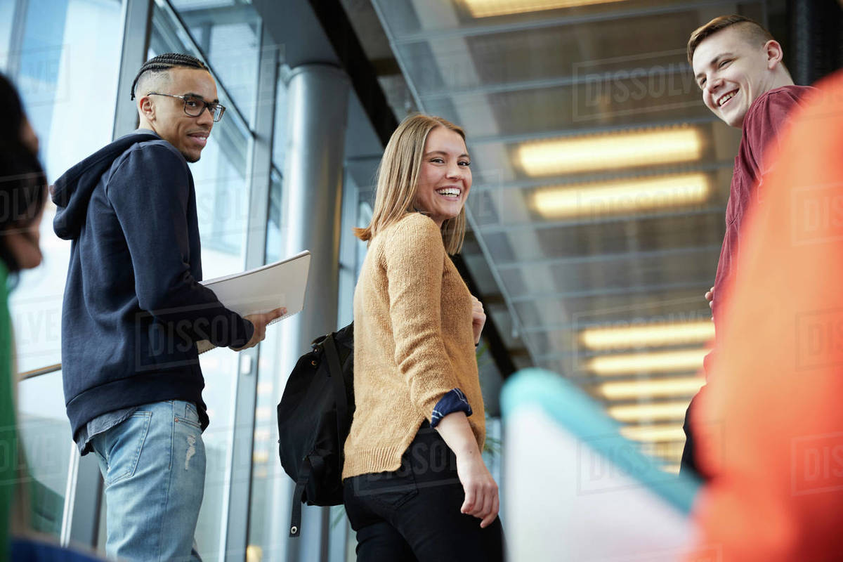 Smiling students looking at friend while standing by window in ...