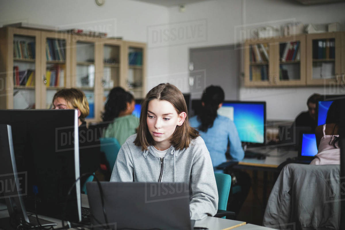 Confident female student using laptop at desk against teacher and ...