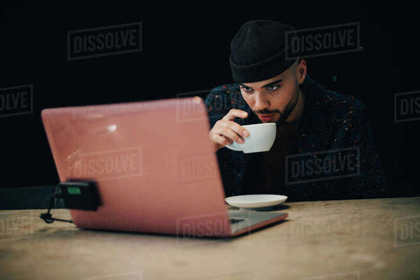 Confident young male hacker drinking coffee while looking at laptop on ...