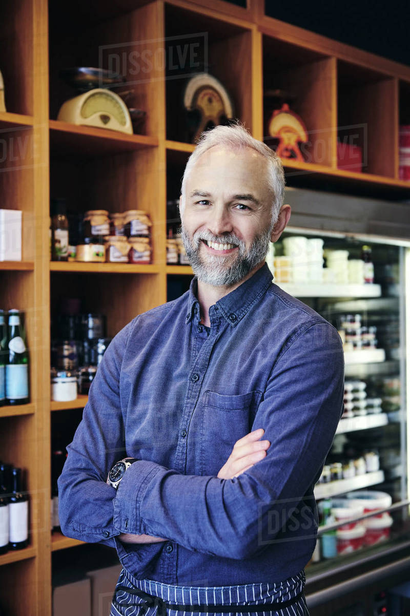 Portrait of confident mature salesman standing by rack in deli ...