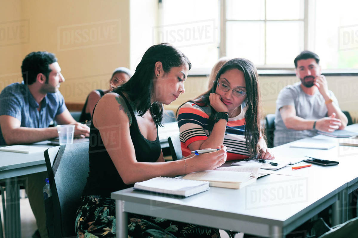 Female students discussing lessons while sitting at desk in classroom ...