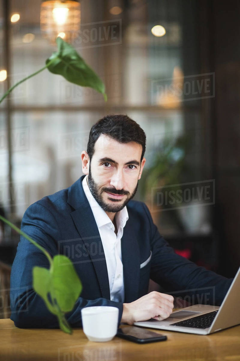 Portrait of confident male entrepreneur using laptop while sitting at ...
