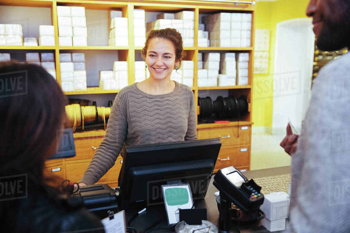 Smiling young female cashier looking at customer standing in furniture ...