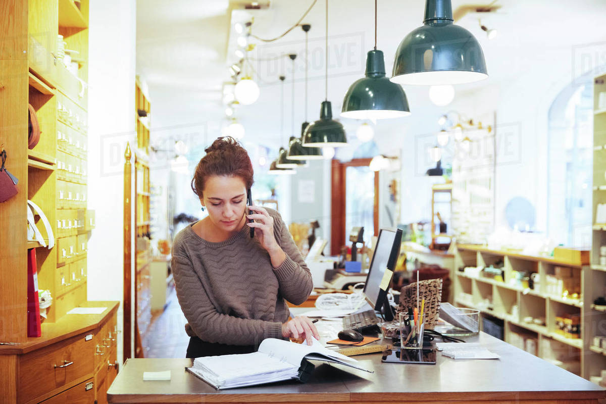 Young female sales clerk talking on mobile phone while looking at file ...