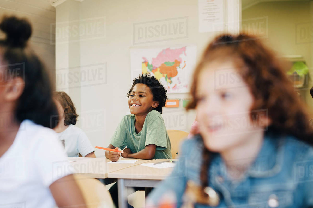 Smiling boy sitting with friends at desk in classroom - Royalty-free ...