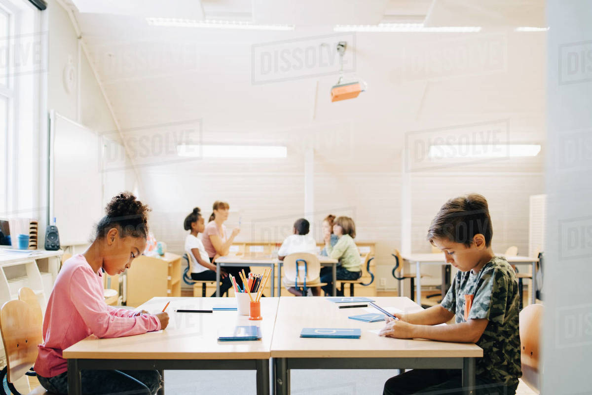Boy and girl writing at desk while friends learning with student in ...