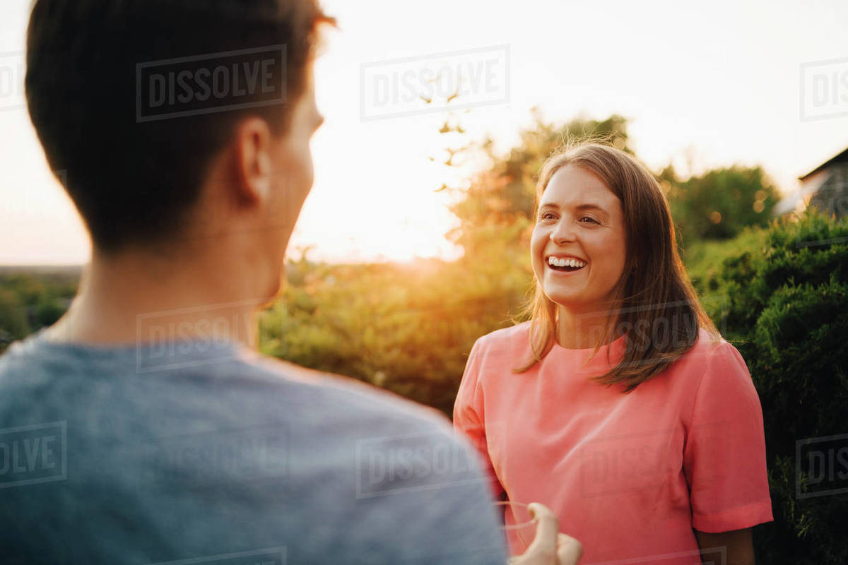 Smiling couple talking while standing at balcony in yard during sunset ...