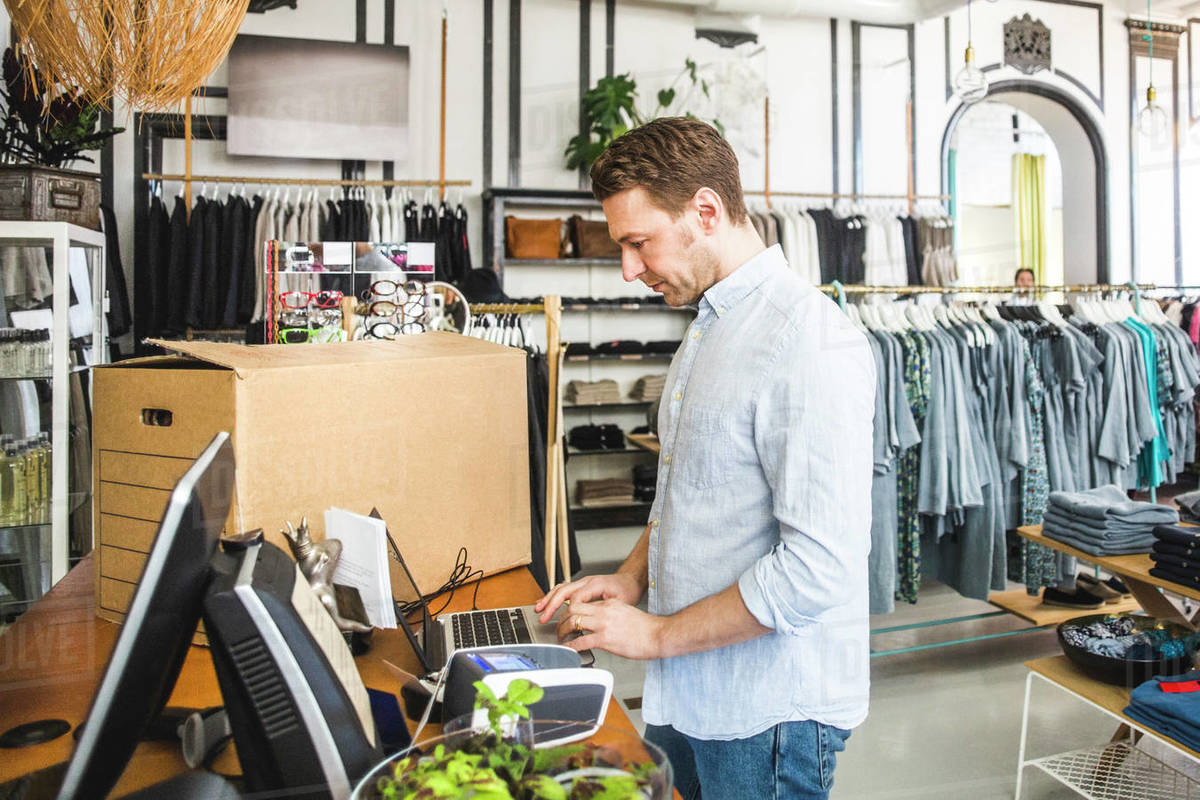 Side view of salesman using laptop while standing in clothing store ...