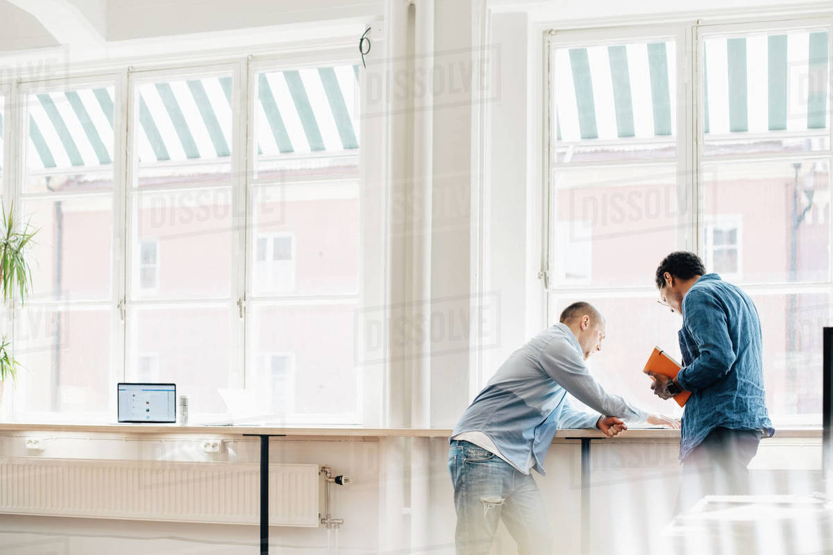 Male computer programmers discussing at desk by window in office ...