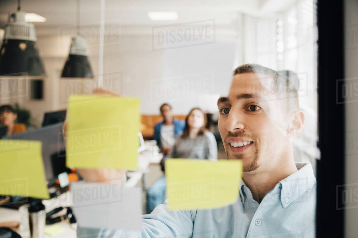 Male computer programmer sticking adhesive note on glass during meeting ...