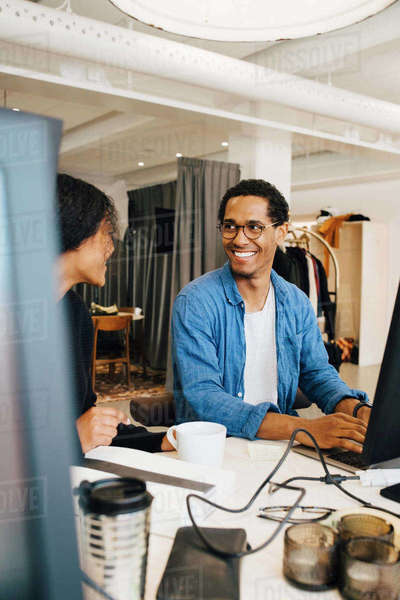 Smiling male and female coworkers talking while sitting at desk in ...