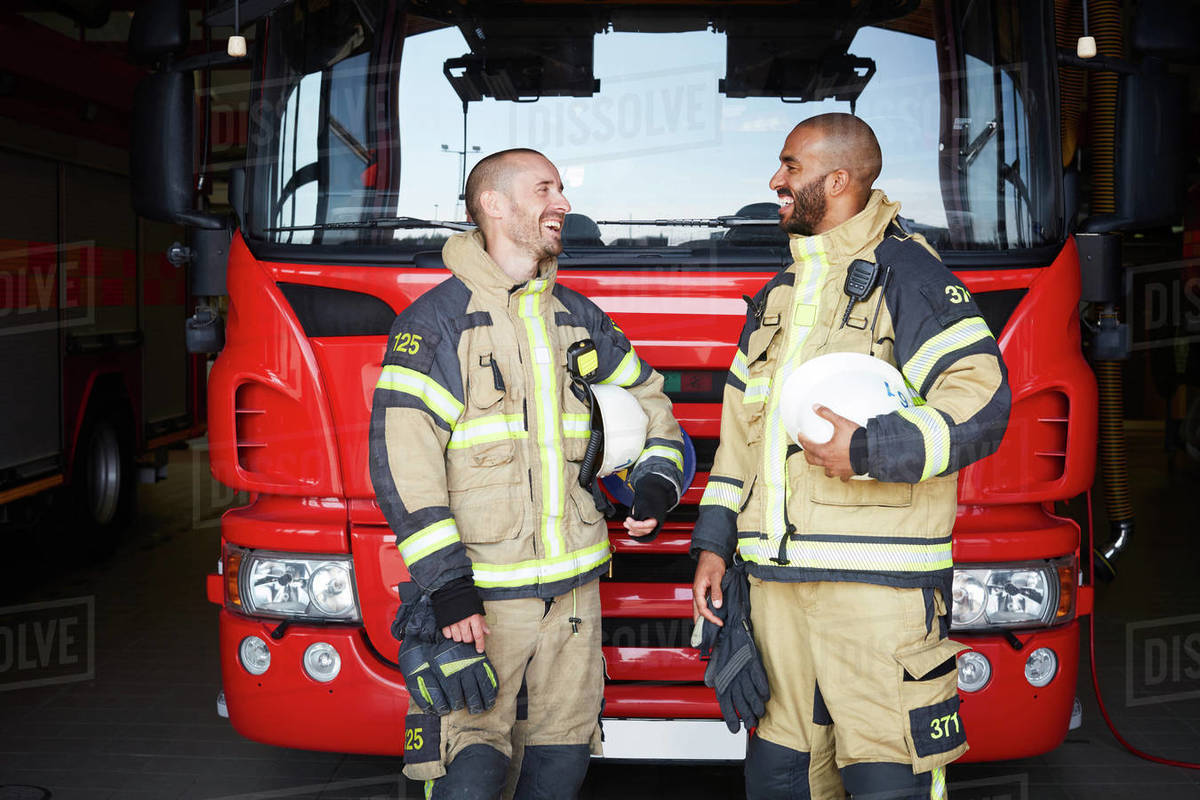 Happy firefighters talking while standing in front of fire engine at ...