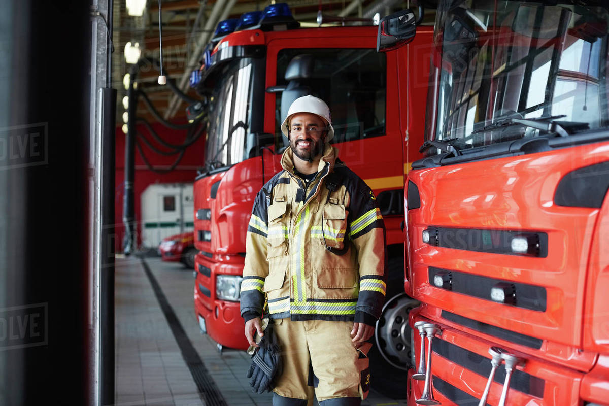 Portrait of smiling firefighter standing by fire engine at fire station ...
