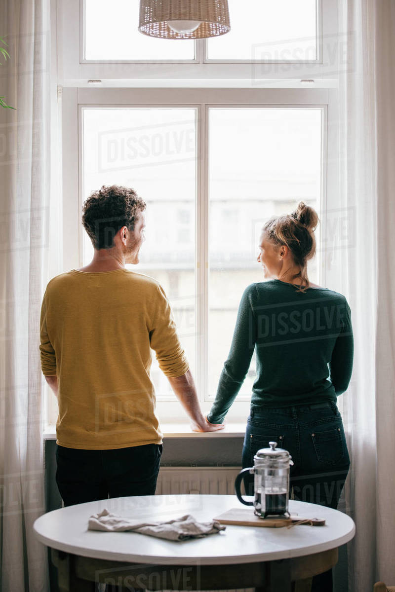 Rear view of romantic couple standing at window in living room ...