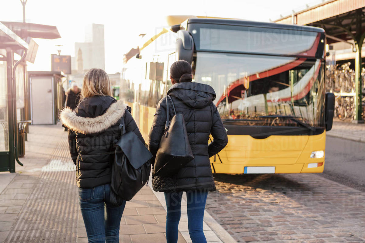Rear view of teenage girls with bags walking on sidewalk by bus in city ...