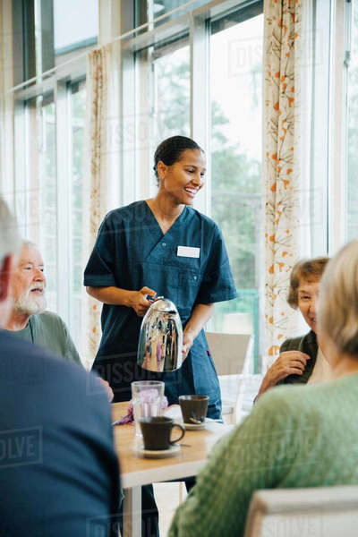 Smiling female nurse serving drink to senior women and men at dining ...