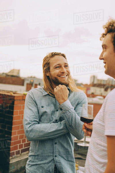 Smiling male friends talking while standing on terrace at rooftop party ...