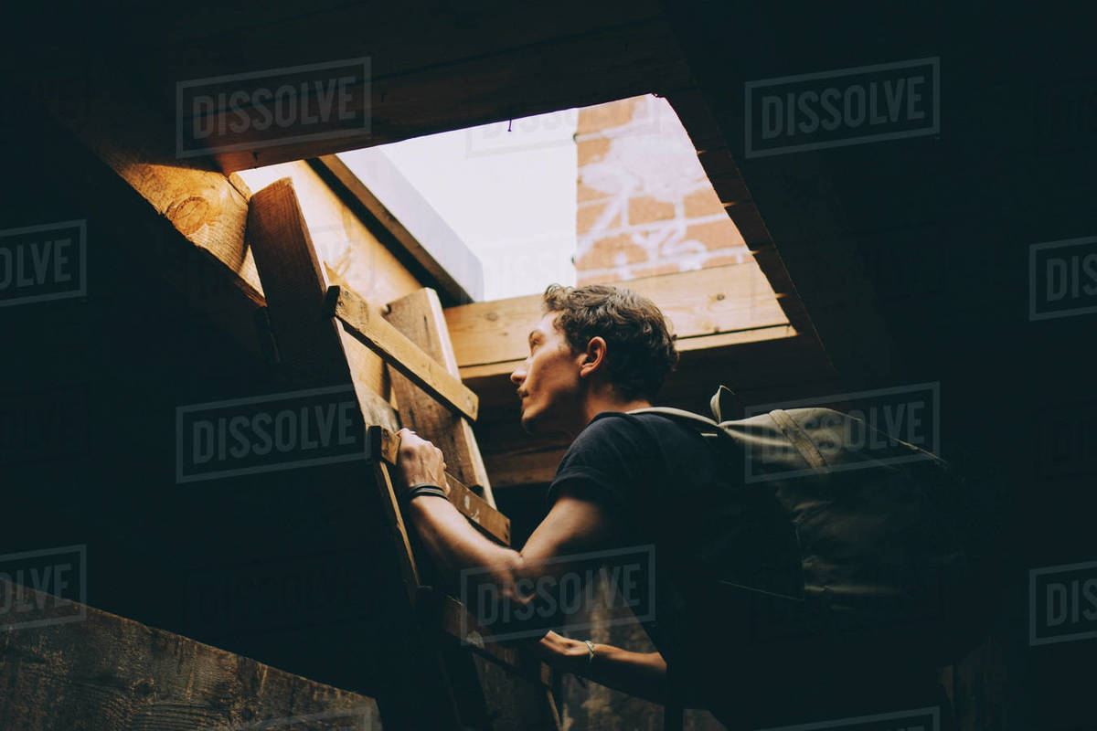 Side view of young man carrying backpack while climbing on ladder ...