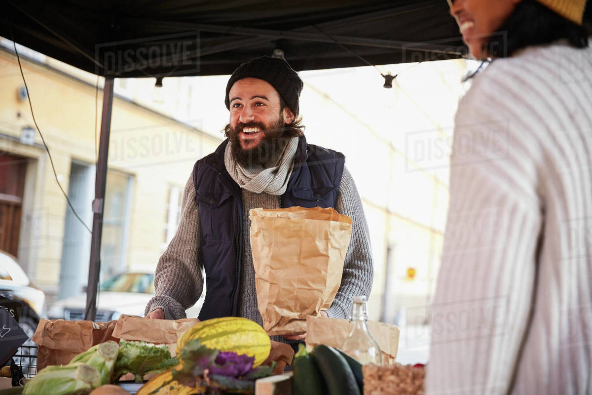 Smiling man buying vegetables from female vendor at market stall ...