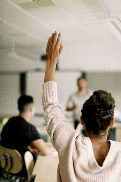 Female student with hand raised while tutor standing in classroom ...