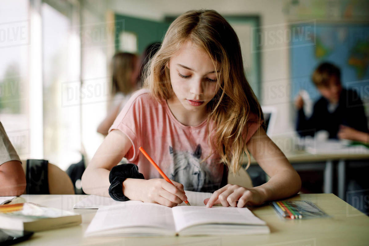 Female student writing in book while sitting at table in classroom ...