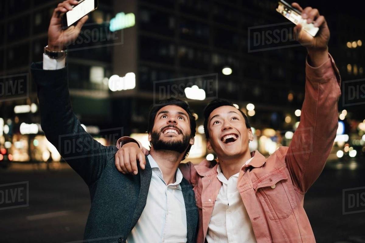 Happy man and male friend looking up with smart phones while gesturing ...