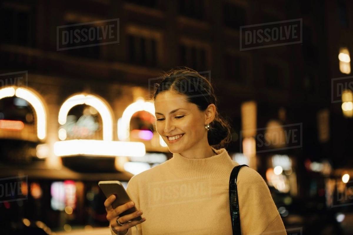 Smiling woman texting through cellphone while standing in city at night ...