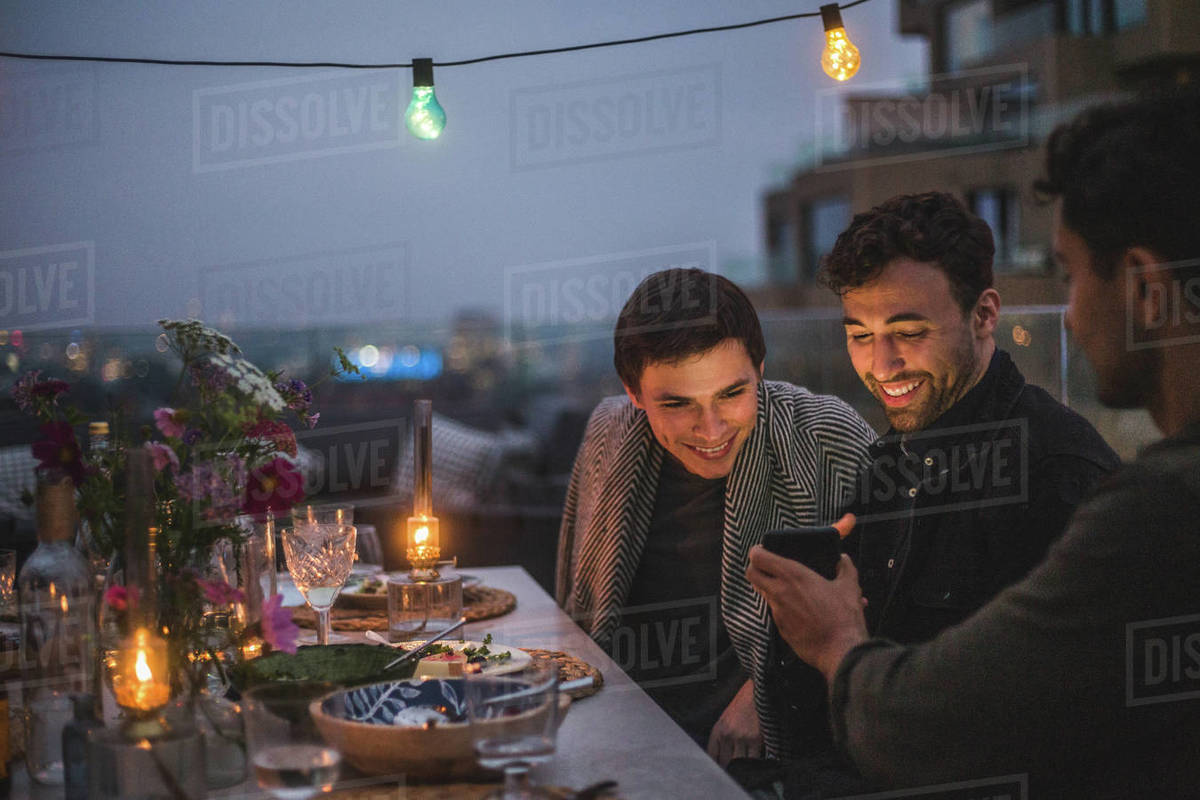 Man showing smart phone to smiling male friends while sitting on ...