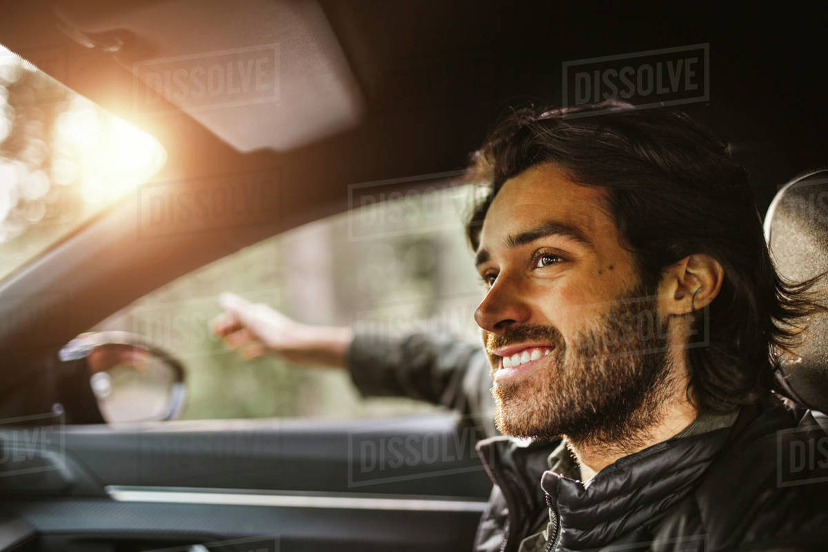 Smiling man with hand outside car window looking away during road trip ...