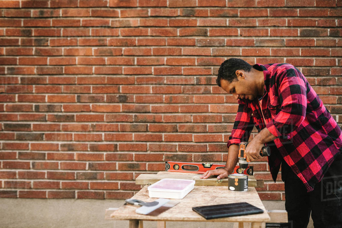Man using drill machine while standing against brick wall during summer ...