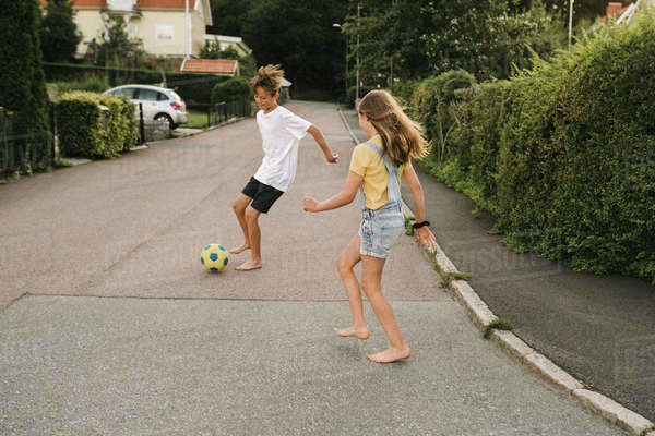 Brother and sister enjoying while playing with soccer ball on road ...