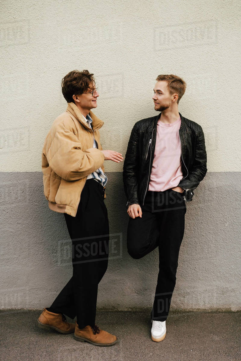 Male friends talking with each other while leaning on beige wall ...