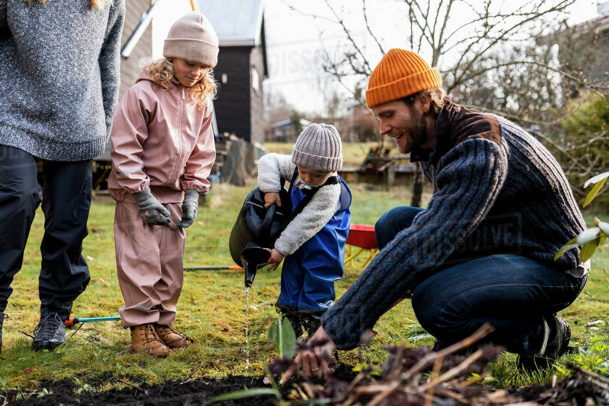 Man guiding son using watering can by girl while doing gardening at ...