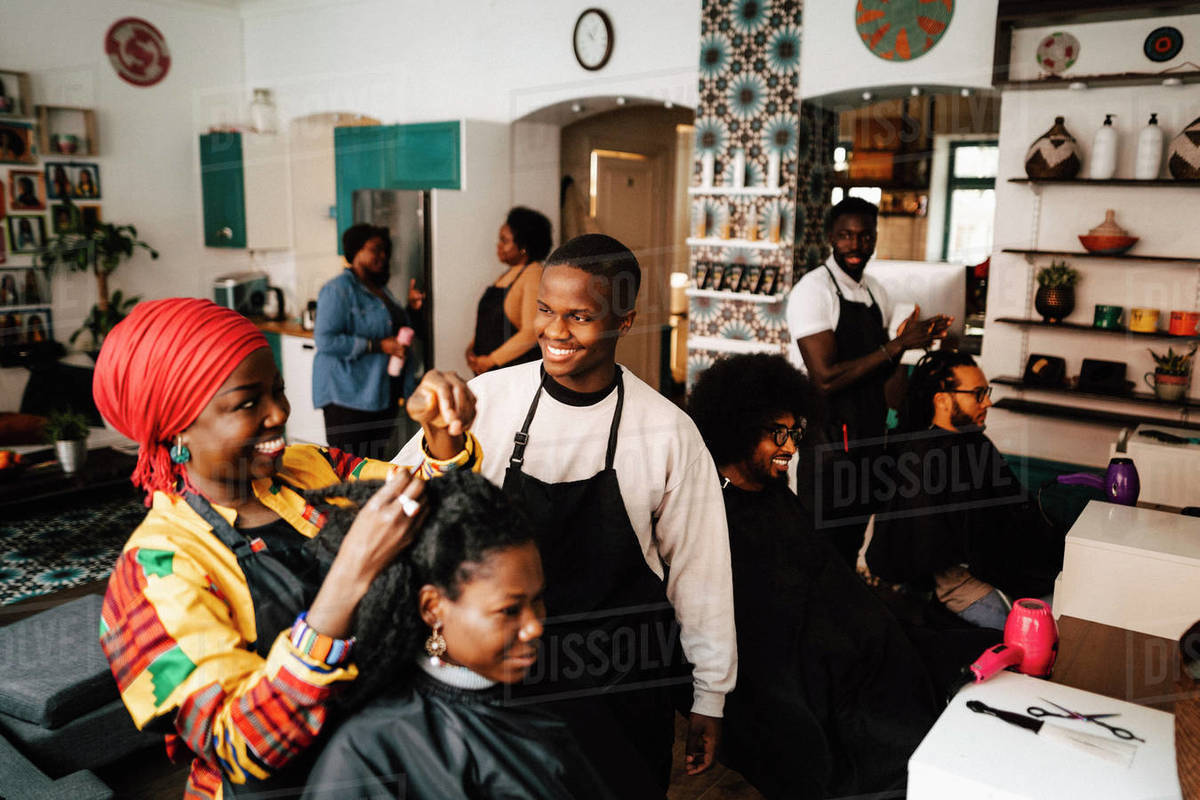 Smiling female hairdresser talking with male coworker while making locs ...