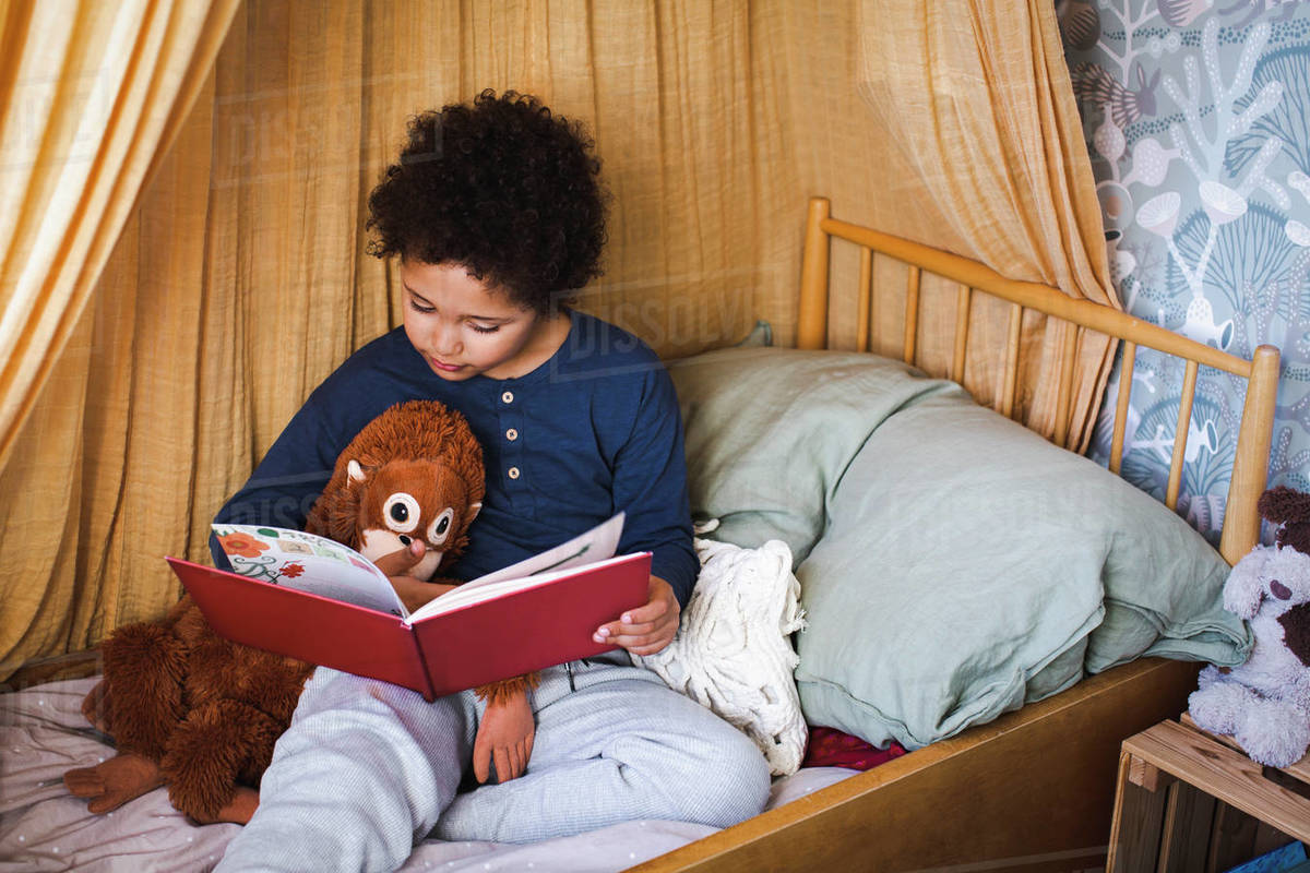 Boy with stuff toy reading story book while sitting on bed at home ...