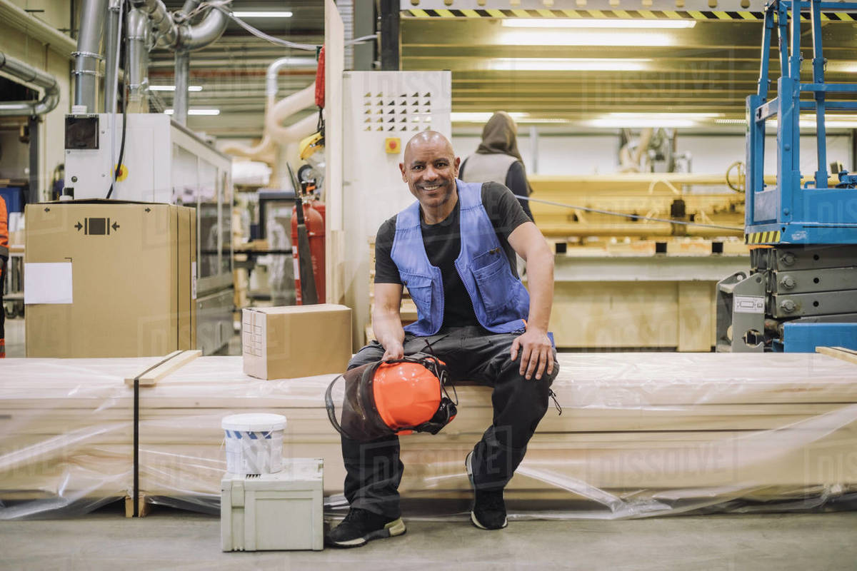 Portrait of happy carpenter sitting with hardhat in warehouse - Royalty ...