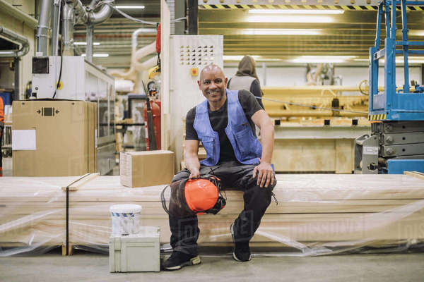 Portrait of happy carpenter sitting with hardhat in warehouse - Royalty ...