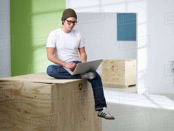Young man sitting on box with laptop - Stock Photo - Dissolve
