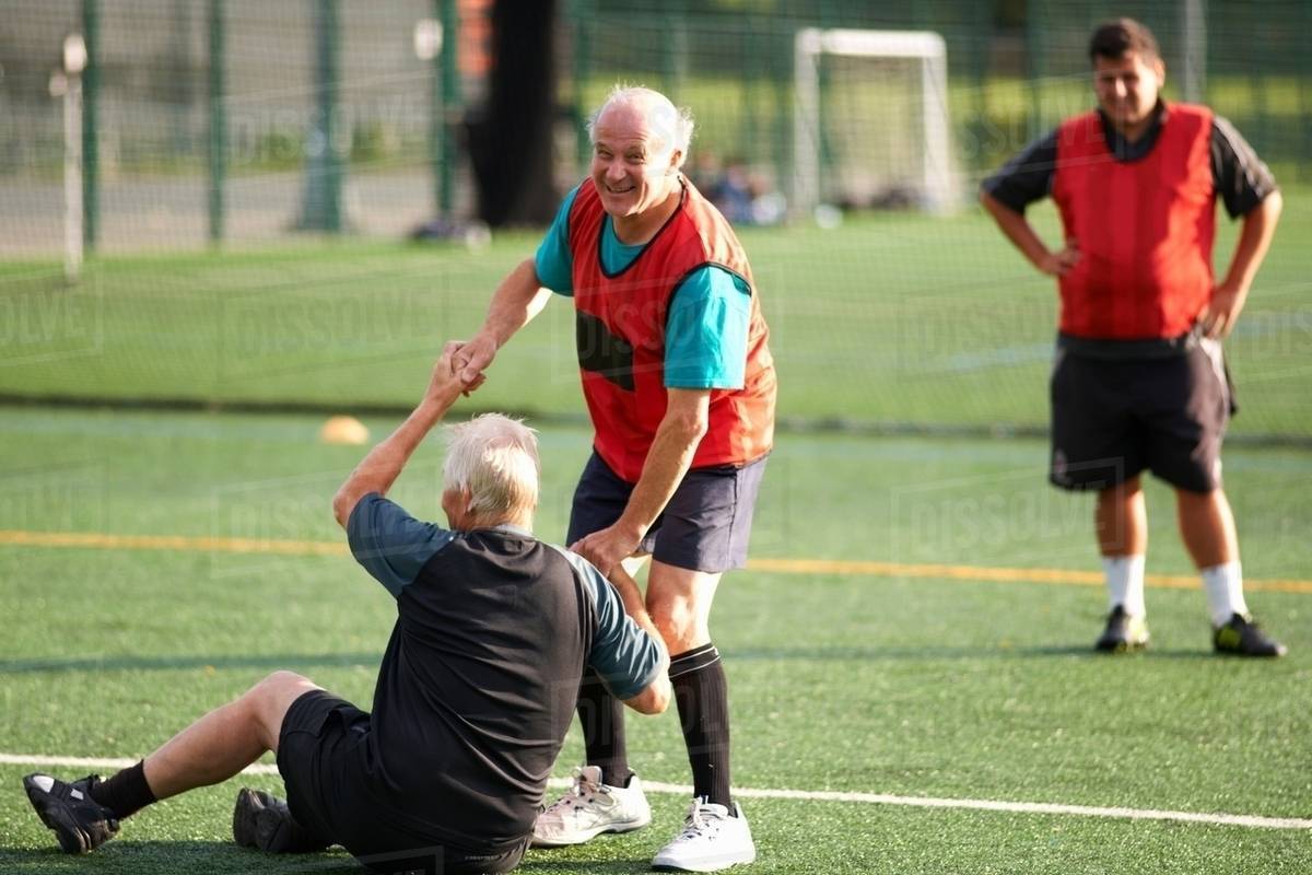 Senior man helping friend stand up on football pitch - Royalty-free ...